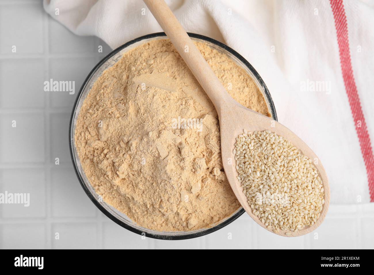 Sesame flour in glass bowl with wooden spoon of seeds on white tiled ...