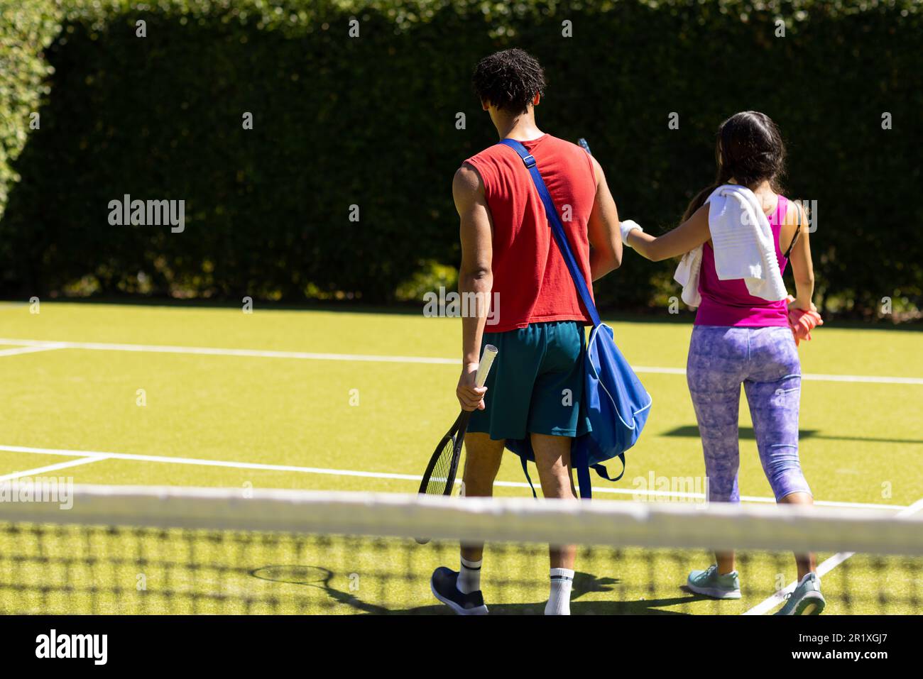 Rear view of diverse couple with bags and rackets walking on sunny ...