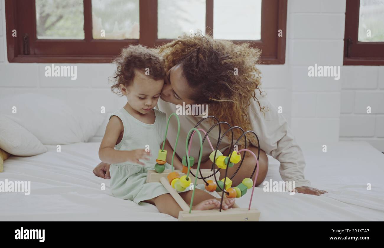 Happy mother and daughter playing with a toy developing colorful wooden blocks on bed together ...
