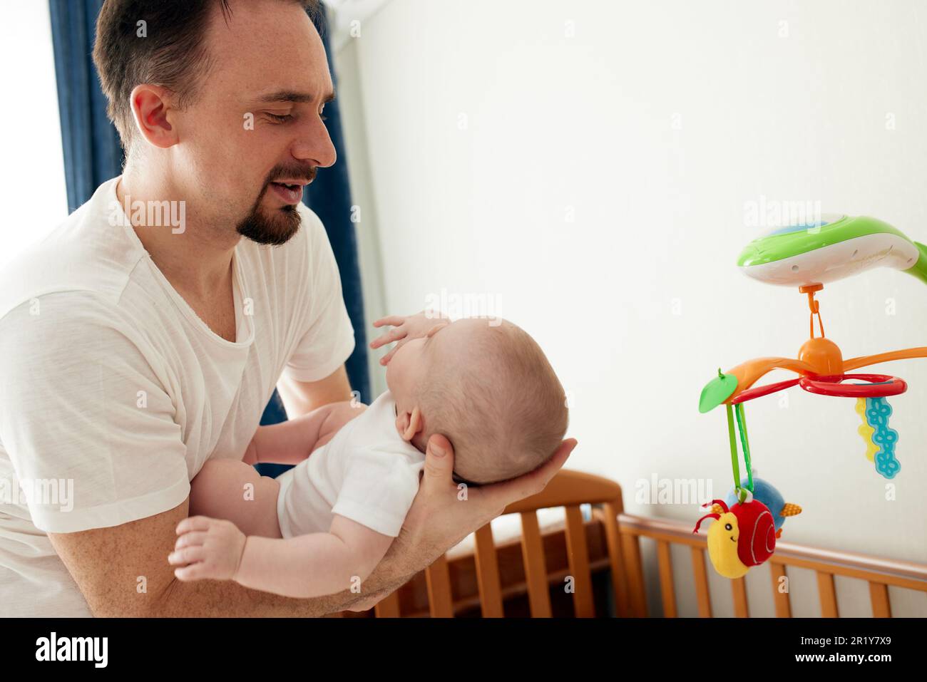 A happy dad puts his baby in a crib with a musical toy. The father hugs ...
