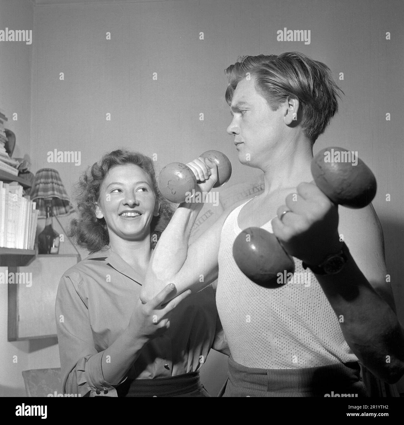 Fun in the 1950s. a man exercises with a pair of dumbbells and his wife ...