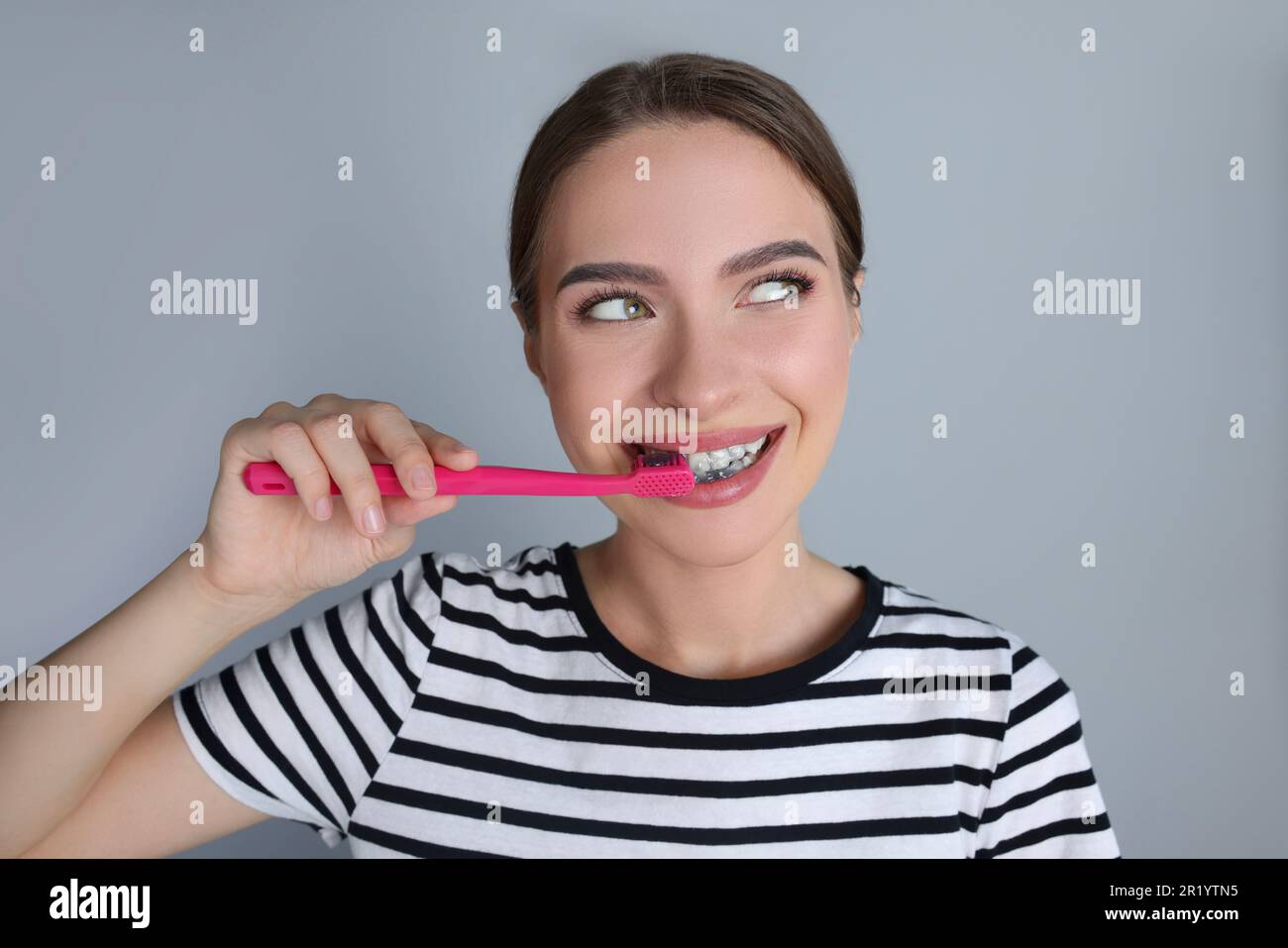 Young woman brushing teeth with charcoal toothpaste on grey background ...