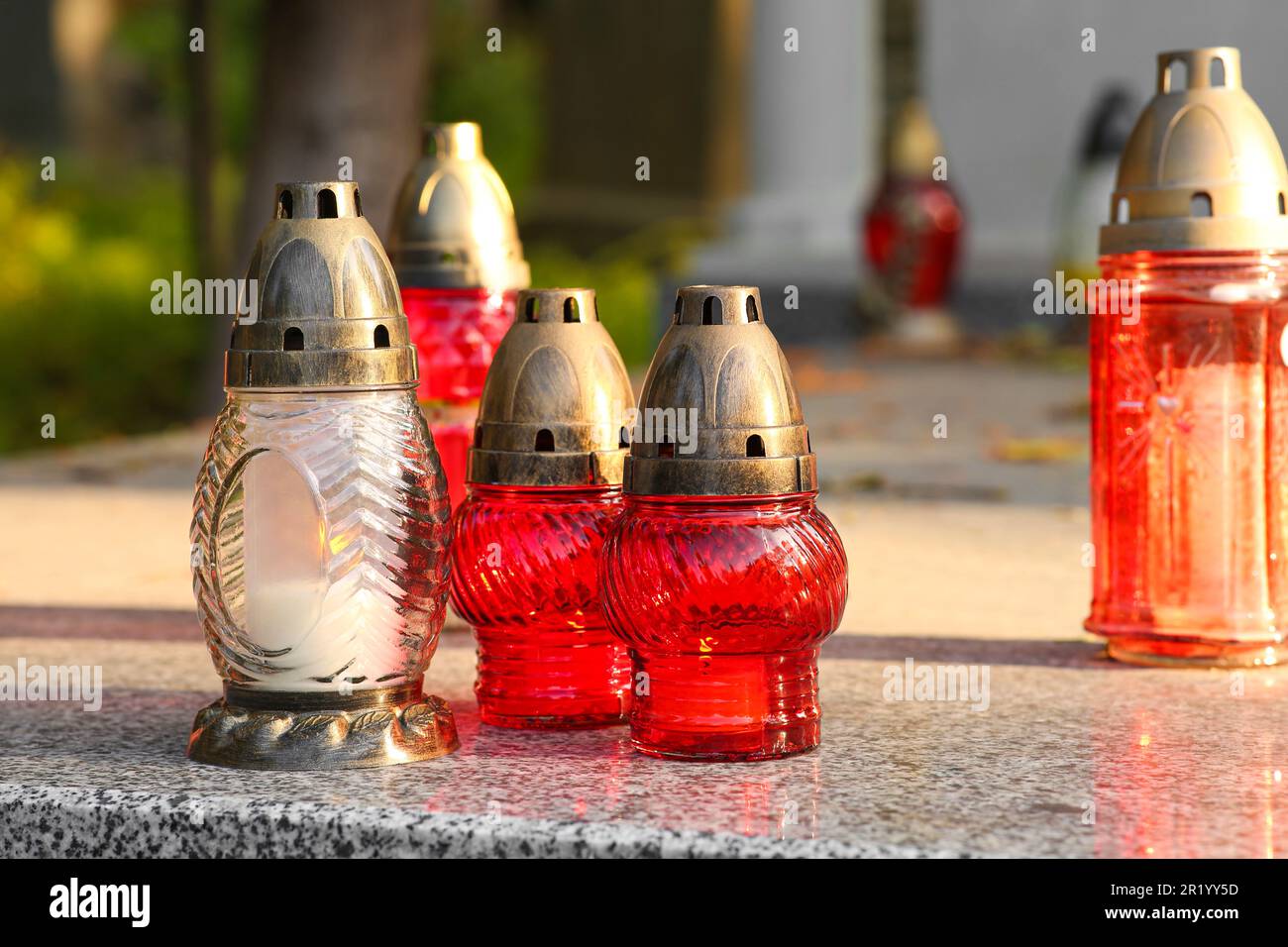 Different grave lanterns with burning candles on granite tombstone outdoors. Funeral ceremony ...