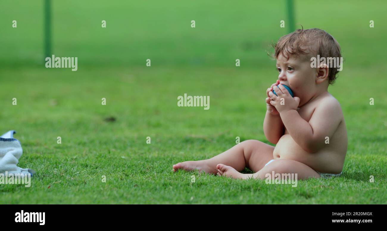 Baby infant seated on grass outdoors with ball inside mouth Stock Photo ...