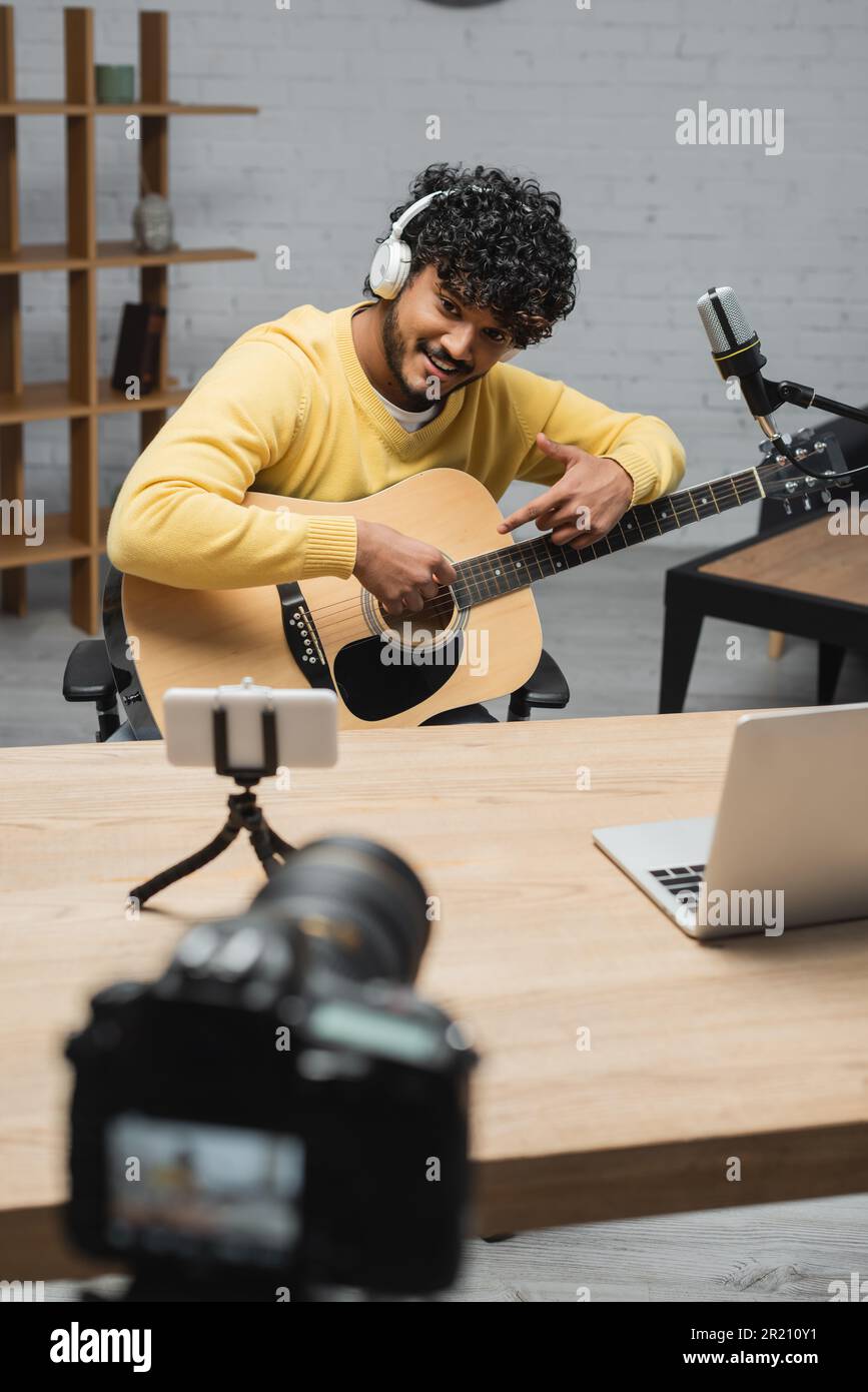 smiling indian musician in headphones pointing at acoustic guitar while ...