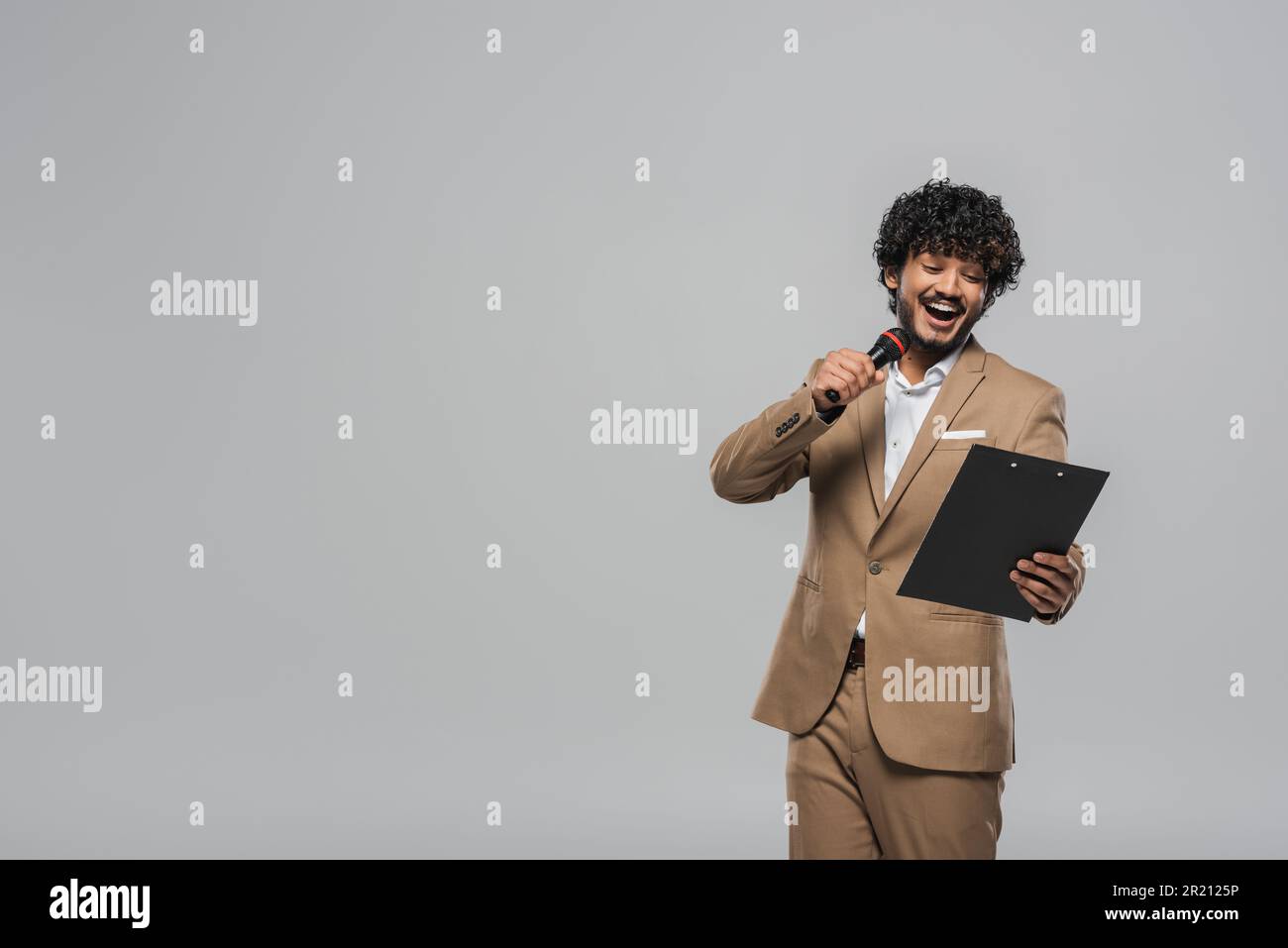 Young and overjoyed indian event host in suit talking at microphone and ...