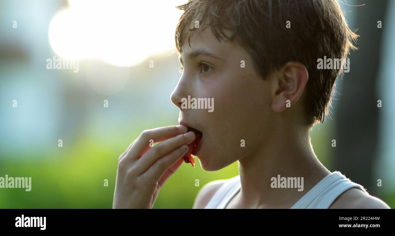Handsome young boy eating fruit and chewing while looking to camera ...