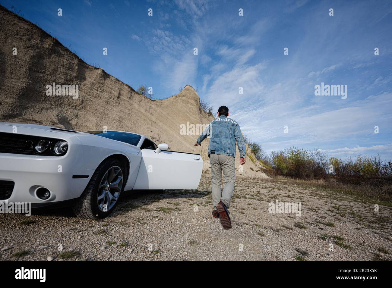 Back of man in jeans jacket and cap open door of his white muscle car ...