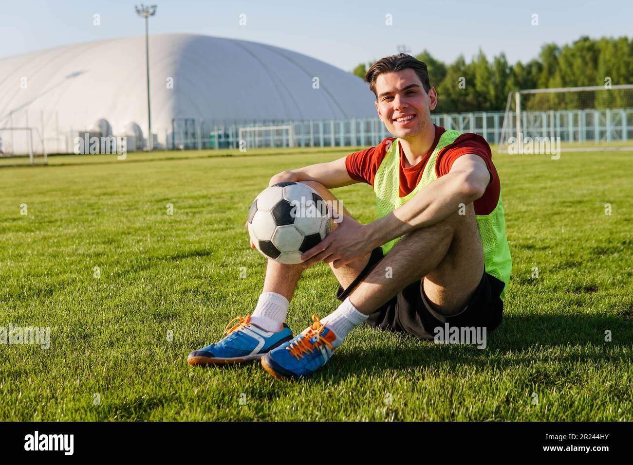 Portrait of a smiling young man football player sitting on field with ...