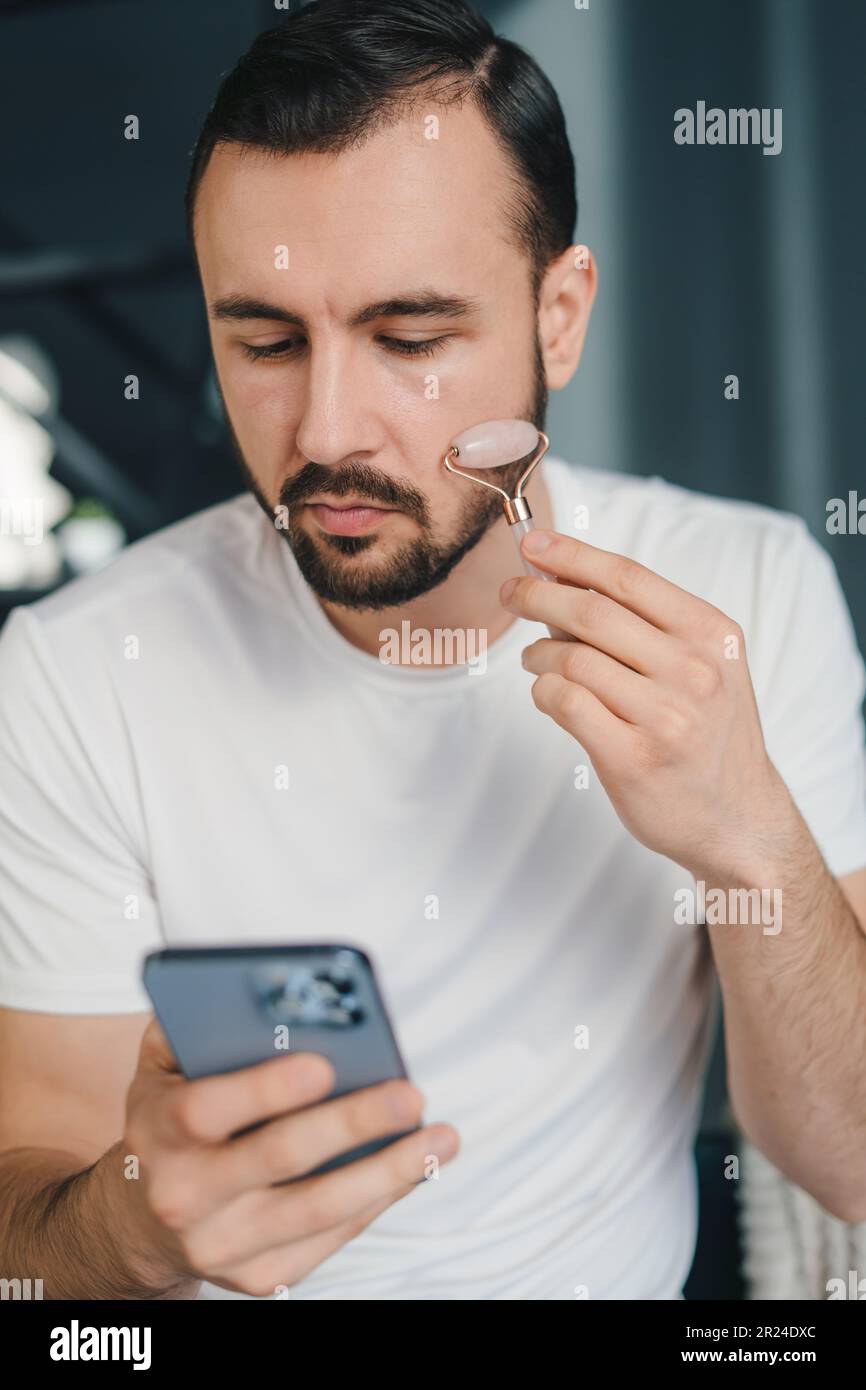 Bearded man using jade face roller at home for beauty facial massage ...