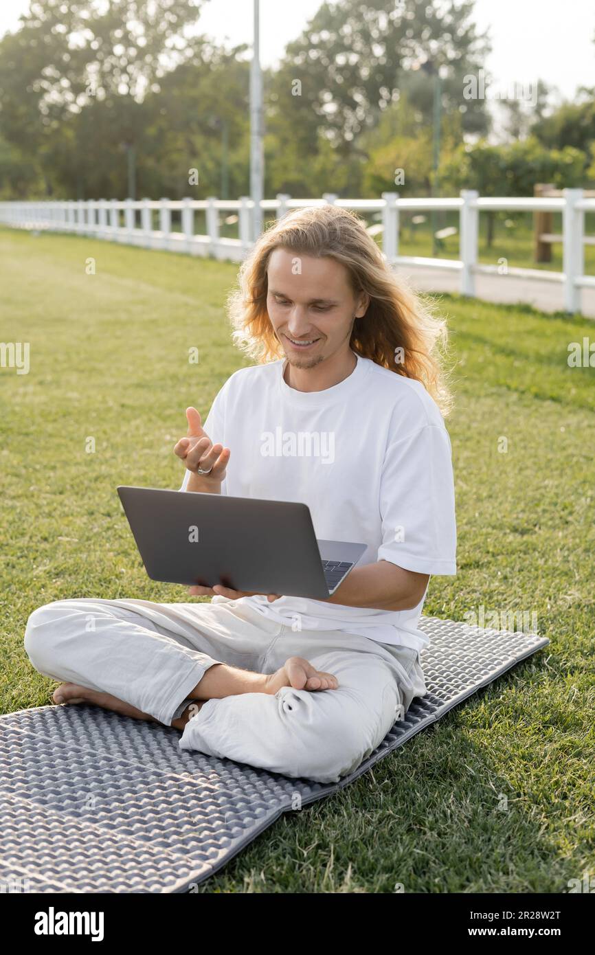 smiling man talking during yoga lesson on laptop while sitting in easy ...