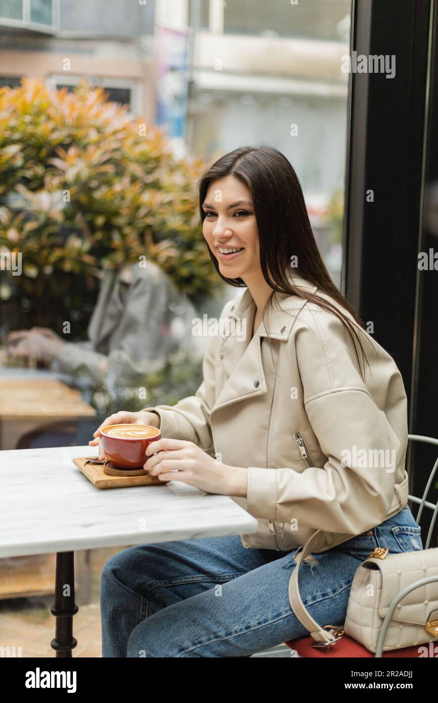 happy brunette woman in leather jacket sitting on chair next to window ...