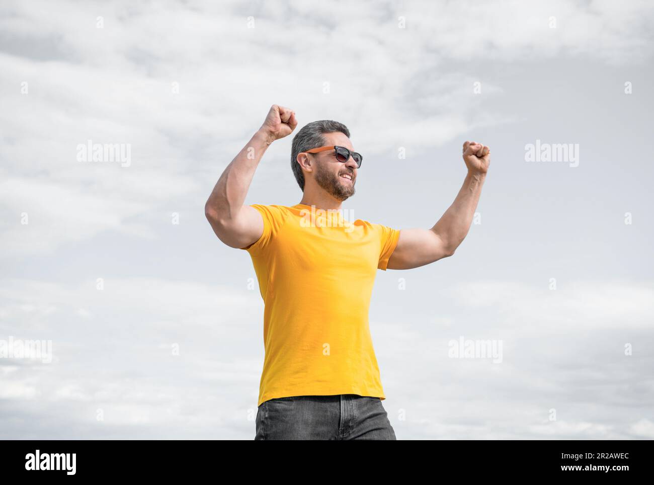 cheerful muscular man in yellow shirt and sunglasses on sky background ...