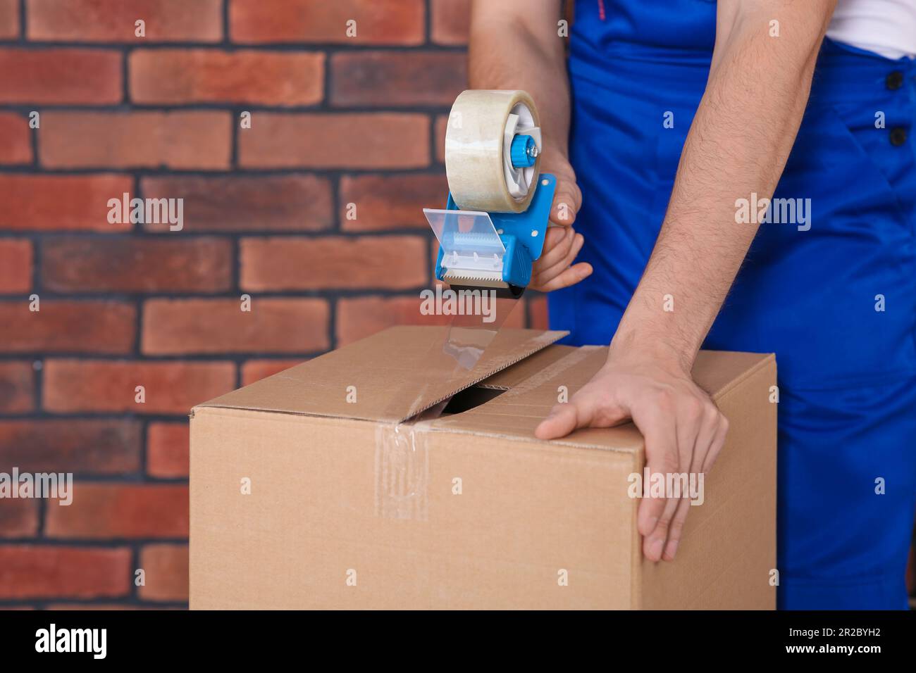 Worker taping box with adhesive tape dispenser near brick wall, closeup ...