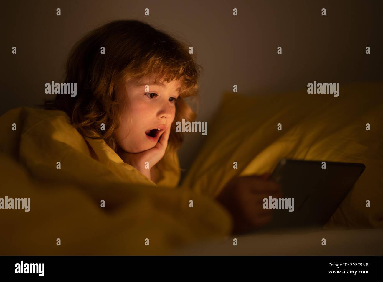 Surprised kid alone at home. Child in bed under a blanket plays on a ...