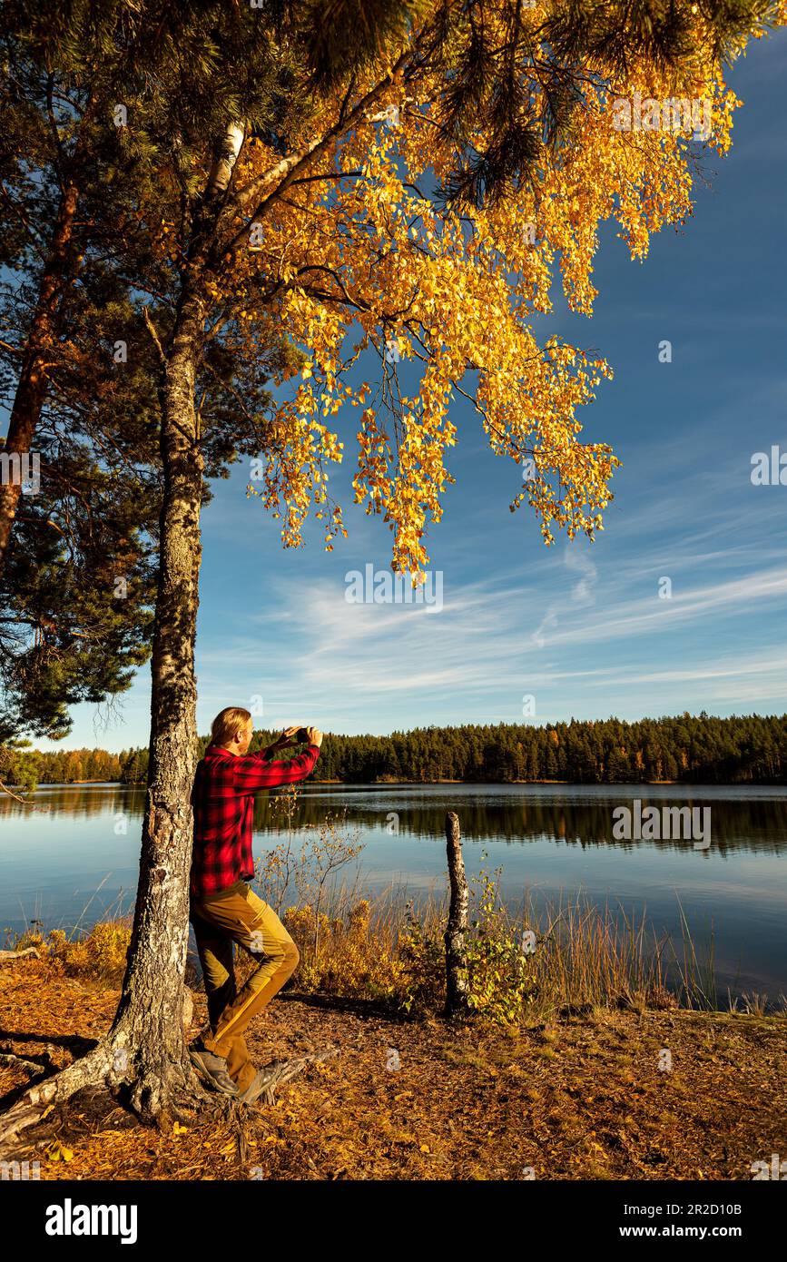 man leaning against tree in colorful fall foliage taking a photo of the ...