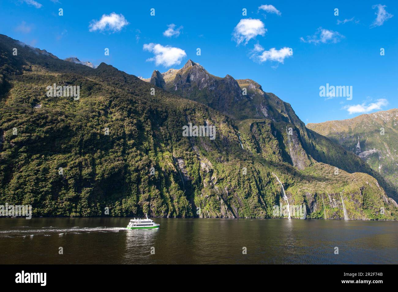 A tour boat glides over smooth water with a backdrop of high, rugged ...
