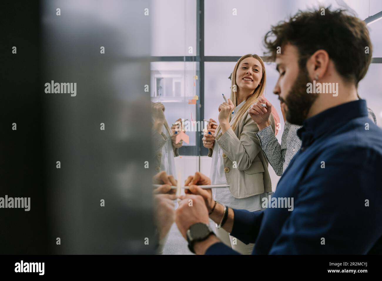 Young smiley blonde woman talking to her colleague while writing ideas ...