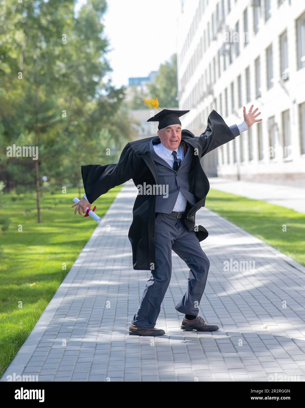 Old happy man in graduation gown jumping outdoors and holding diploma ...