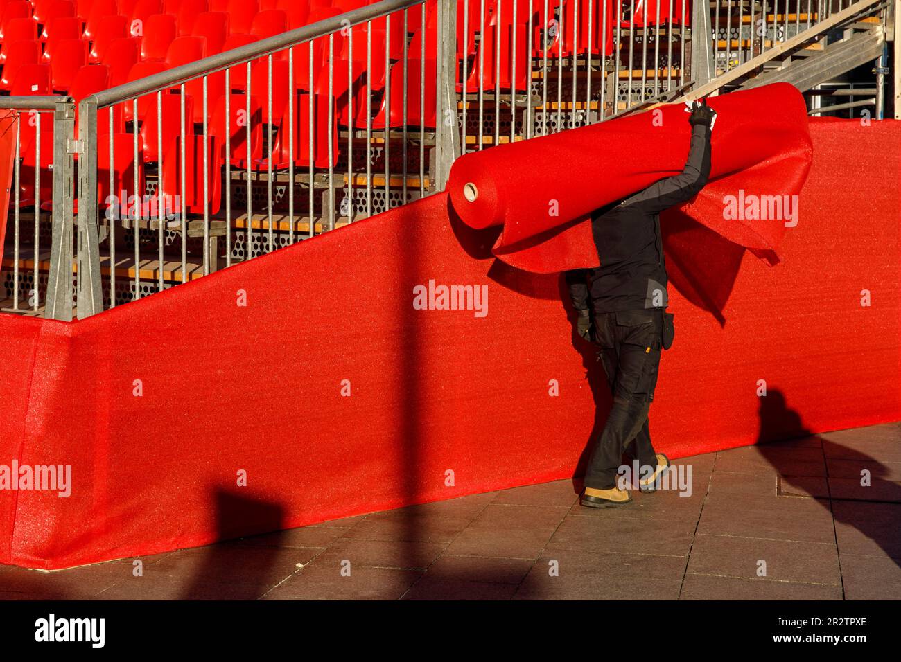 worker carrying a roll of red tarpaulin in front of a red grandstand ...