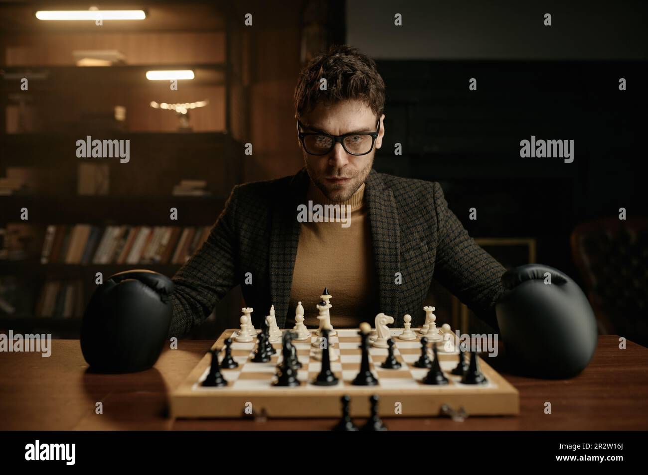 Angry young man wearing boxer gloves sitting at table with chessboard ...