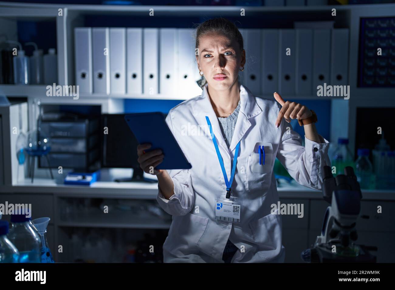 Beautiful blonde woman working at scientist laboratory late at night pointing down looking sad ...