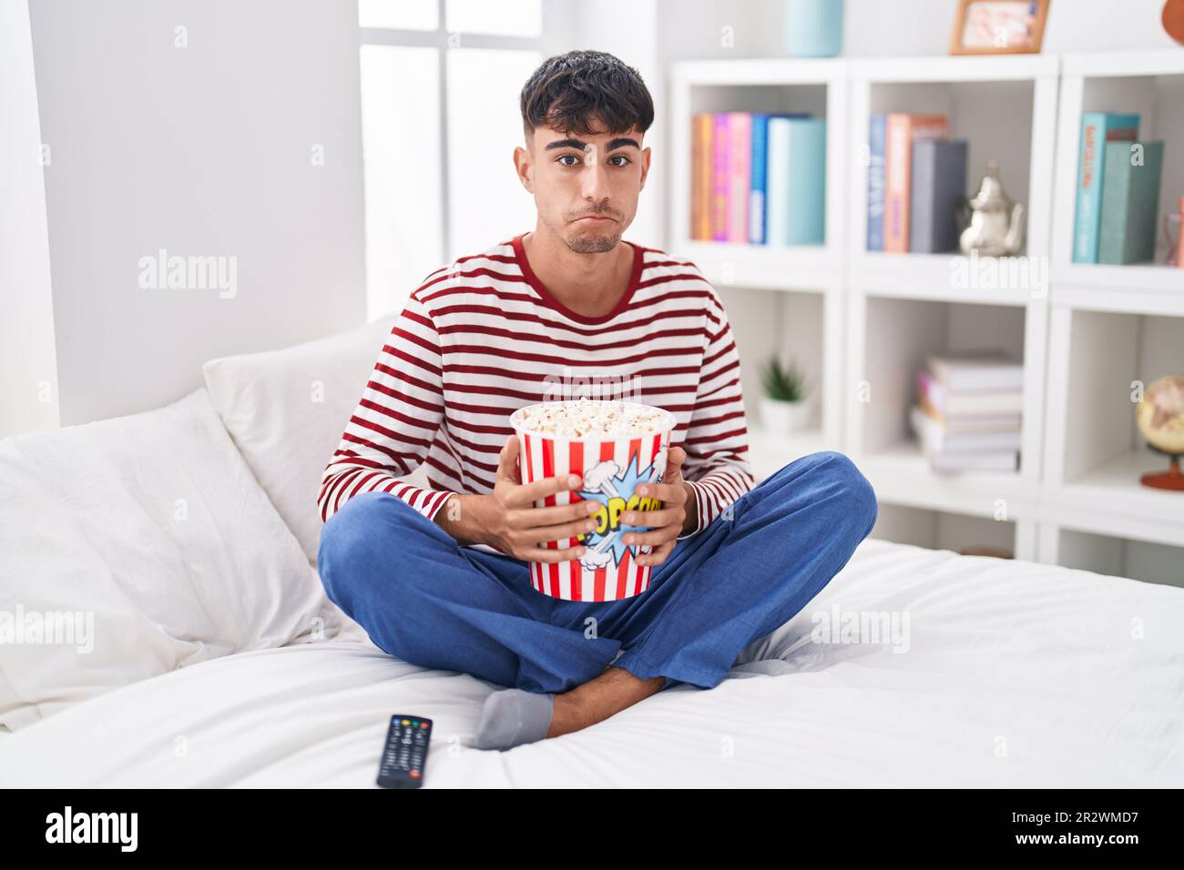 Young hispanic man eating popcorn sitting on the bed watching a movie ...