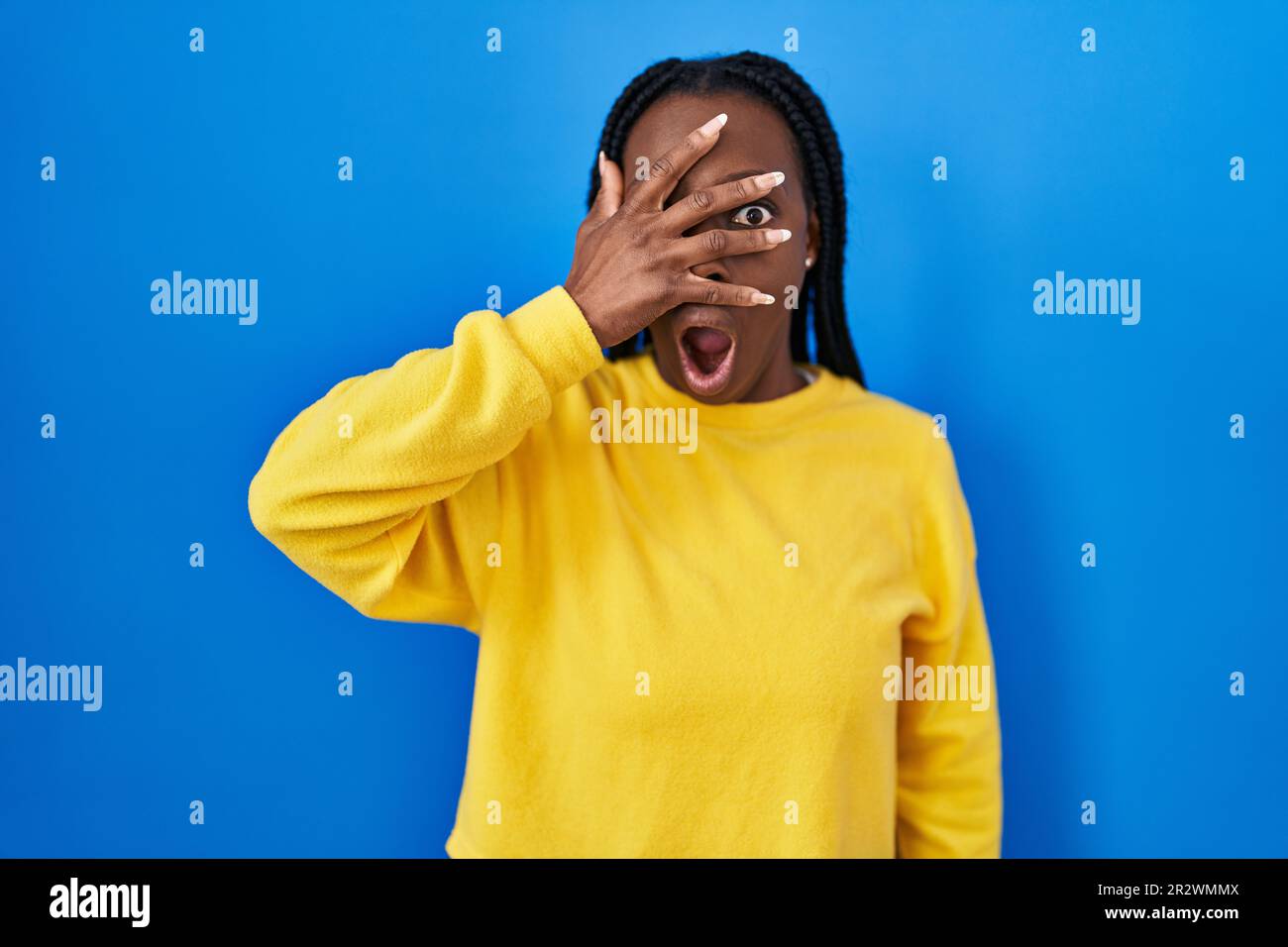 Beautiful black woman standing over blue background peeking in shock ...