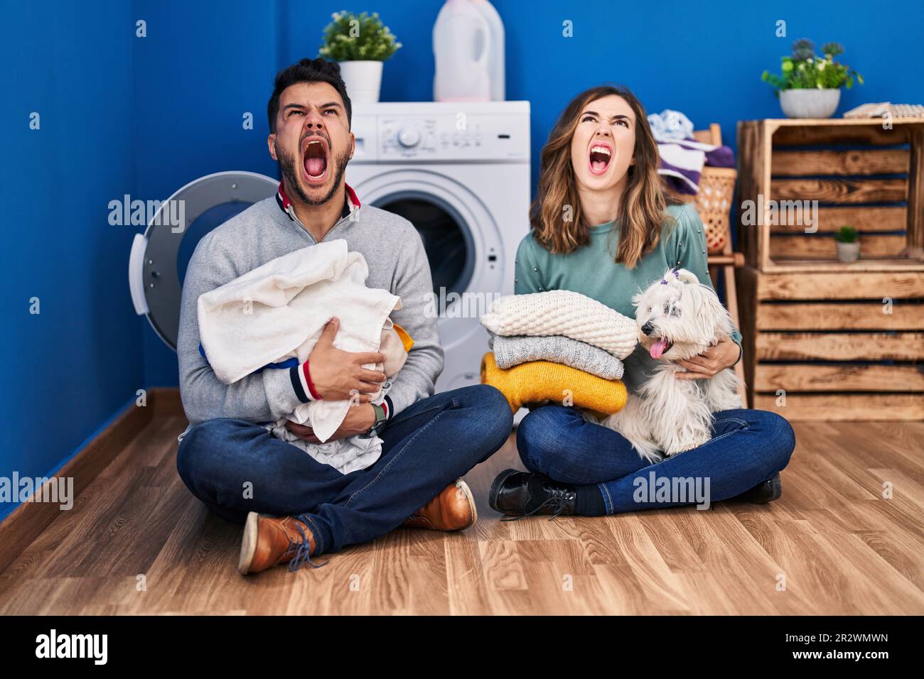 Young hispanic couple doing laundry sitting on the floor angry and mad screaming frustrated and ...