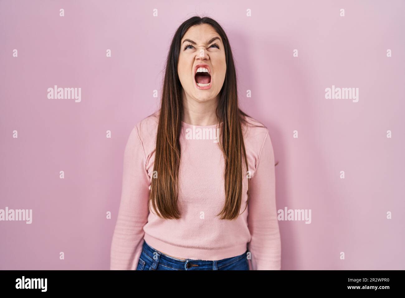 Young brunette woman standing over pink background angry and mad ...