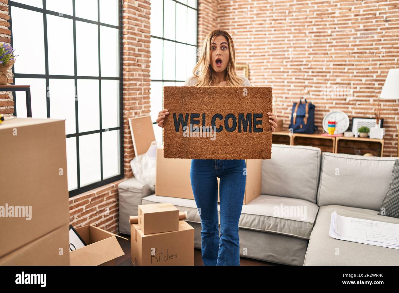 Young blonde woman holding welcome doormat at new home afraid and ...