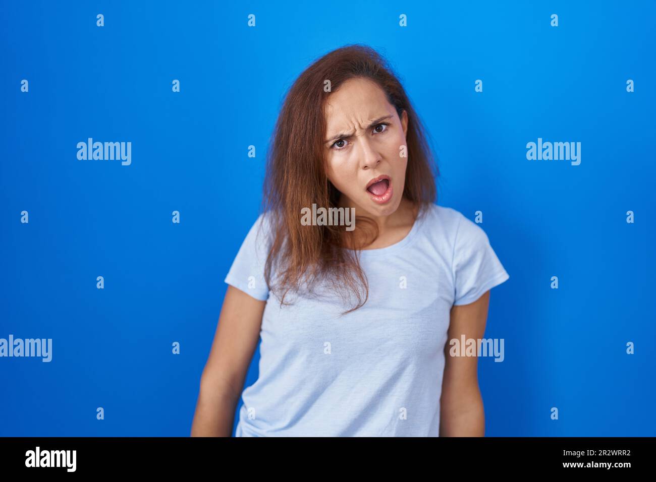 Brunette woman standing over blue background in shock face, looking ...