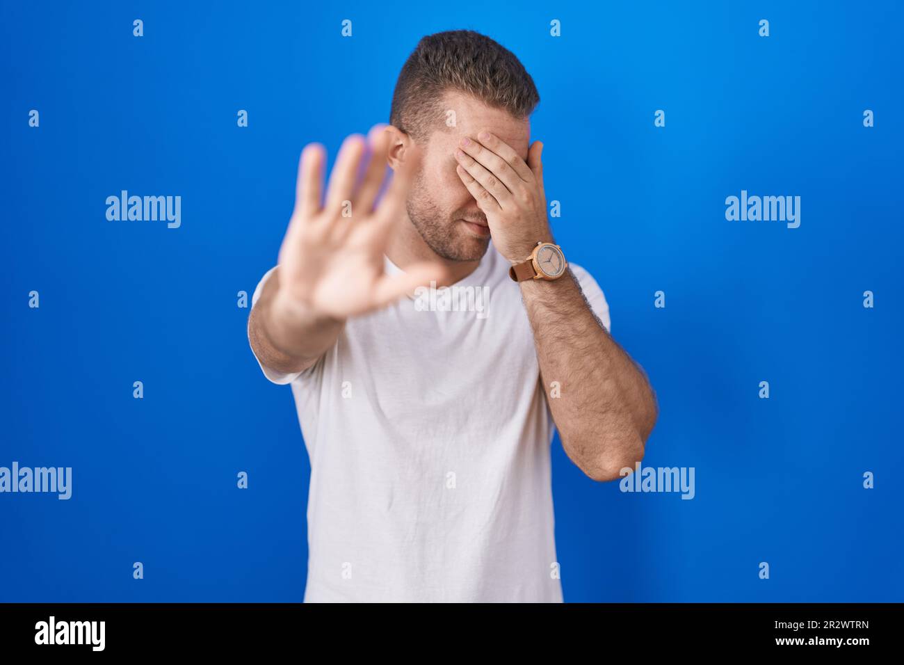 Young caucasian man standing over blue background covering eyes with ...