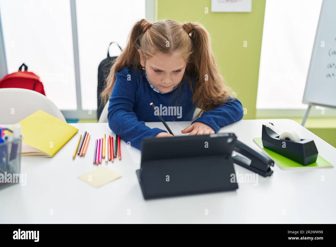 Adorable blonde girl student using touchpad writing notes at classroom ...