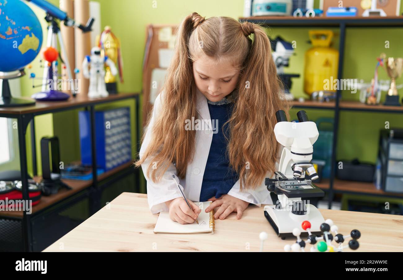 Adorable blonde girl student writing notes at laboratory classroom ...
