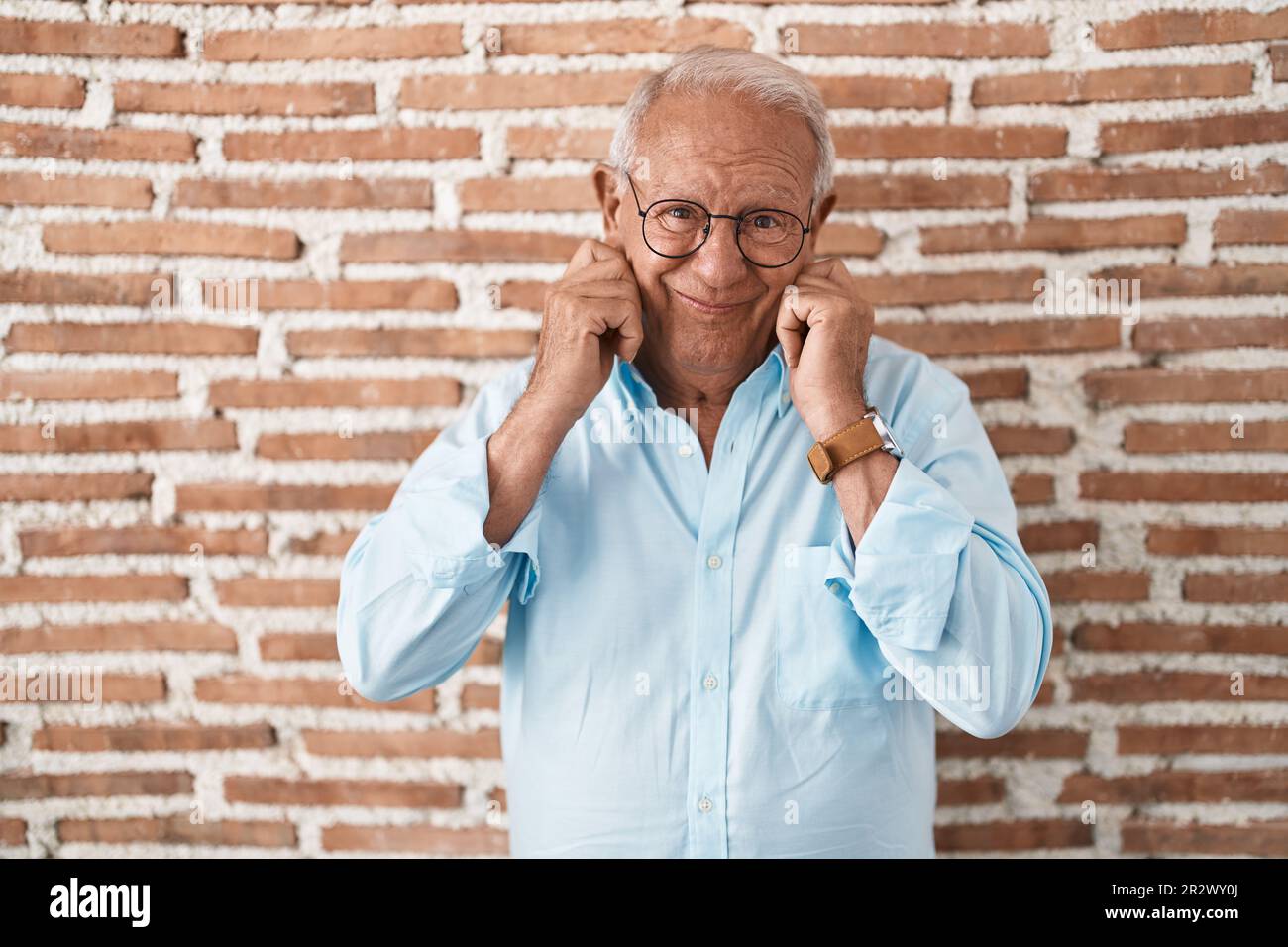 Senior man with grey hair standing over bricks wall covering ears with ...