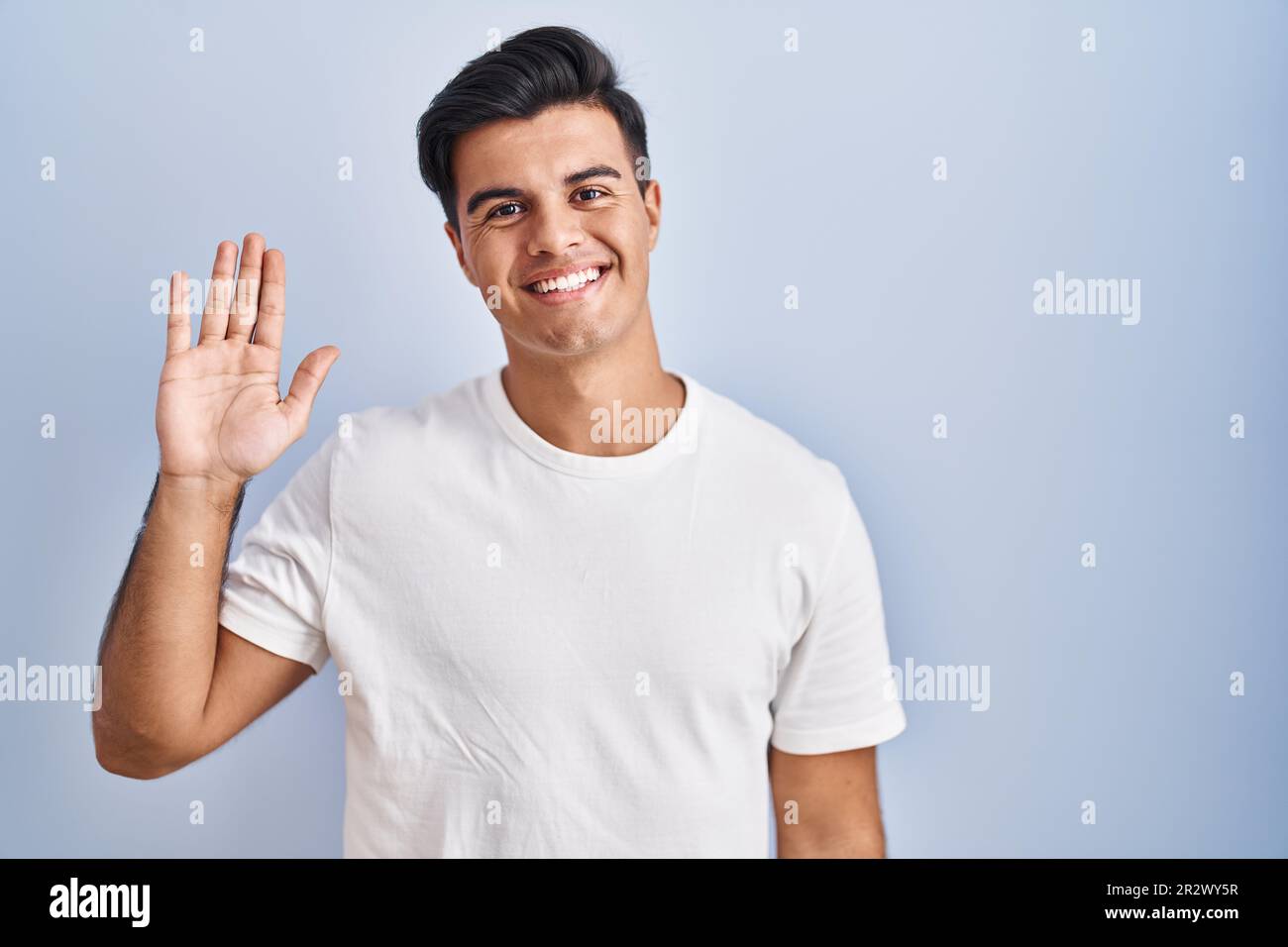 Hispanic man standing over blue background waiving saying hello happy ...