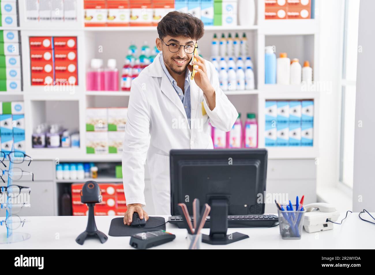 Young arab man pharmacist using computer talking on smartphone at ...