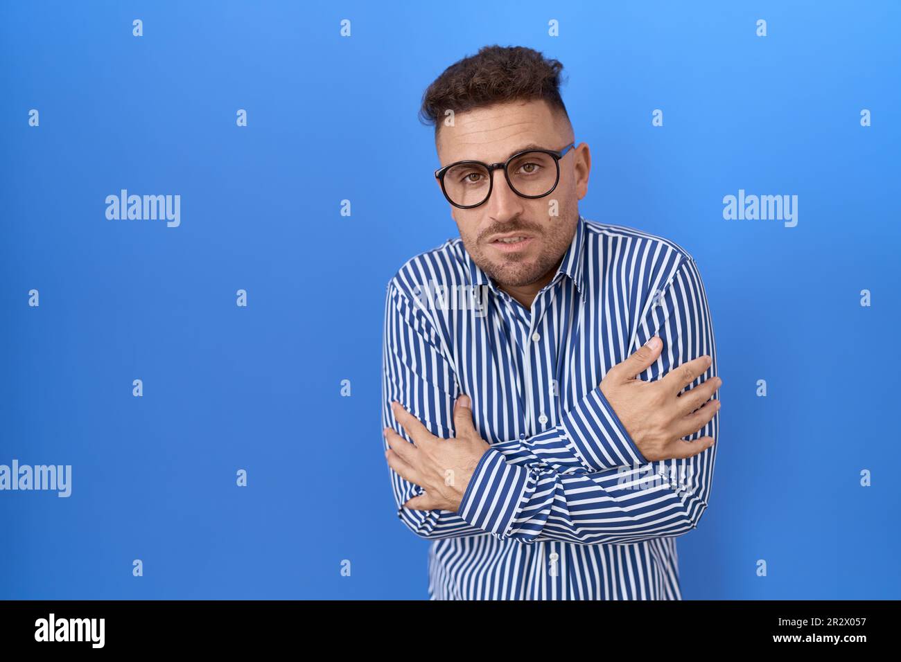 Hispanic man with beard wearing glasses shaking and freezing for winter ...