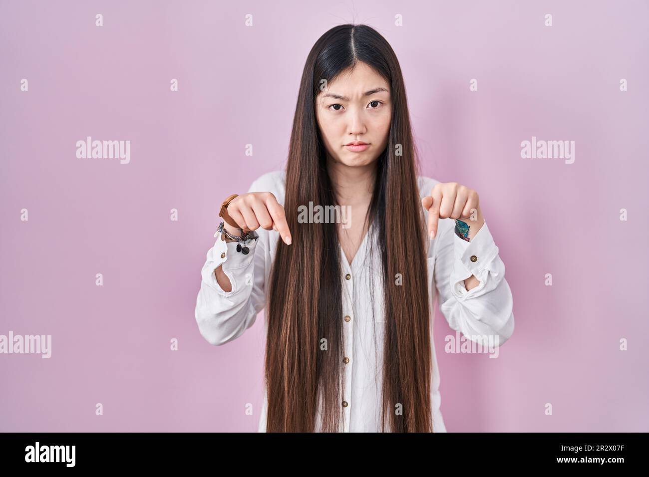 Chinese young woman standing over pink background pointing down looking ...