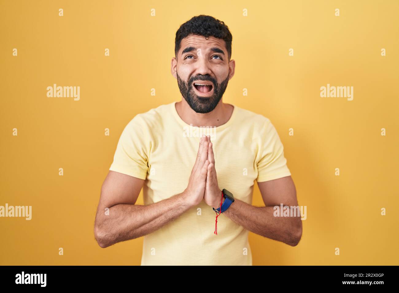 Hispanic man with beard standing over yellow background begging and ...