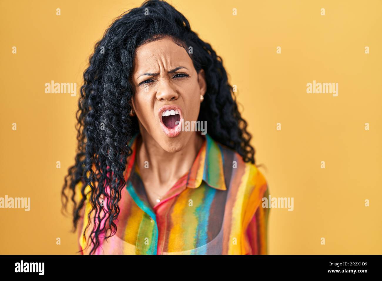 Middle age hispanic woman standing over yellow background angry and mad ...