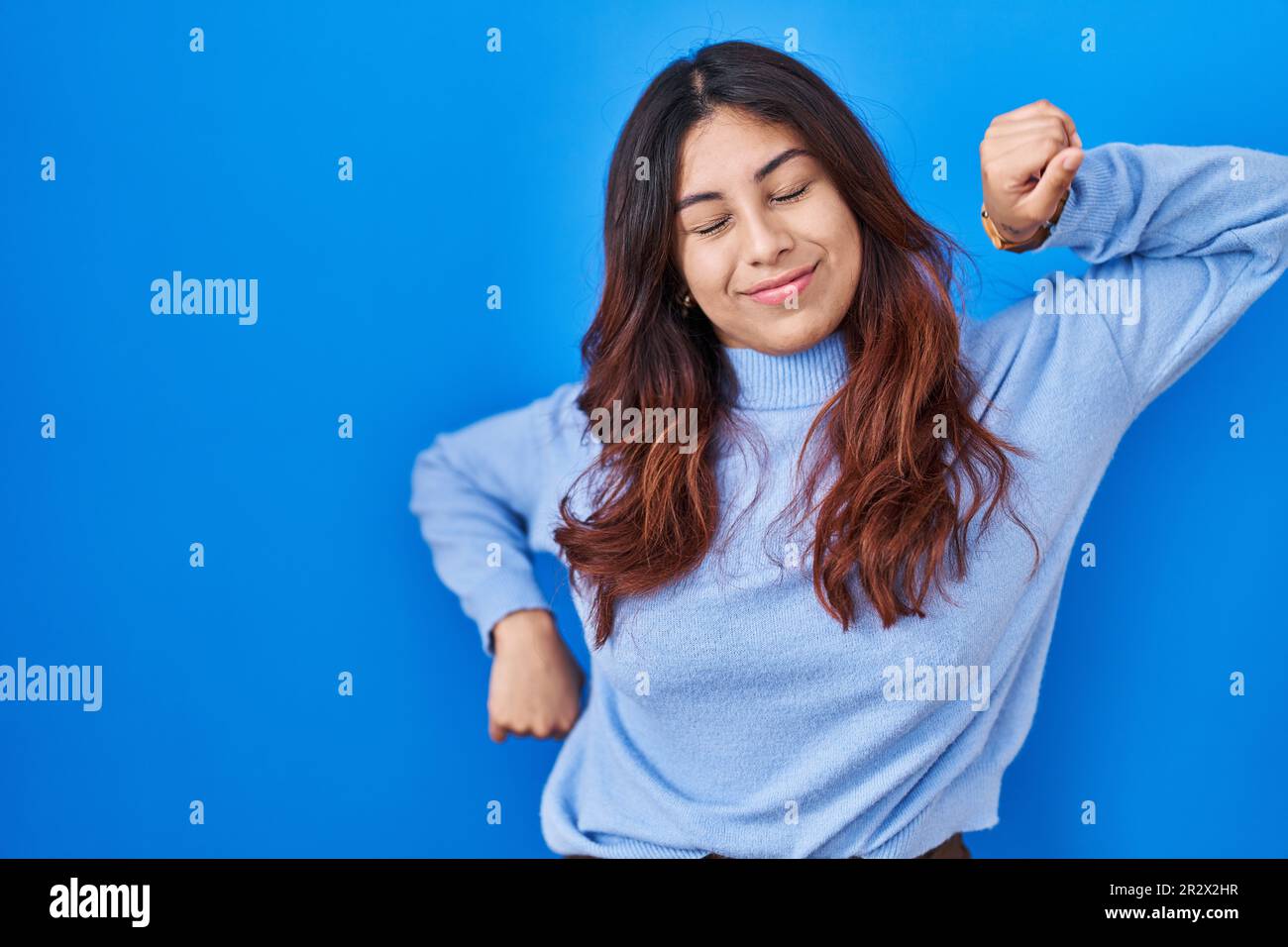 Hispanic young woman standing over blue background stretching back ...