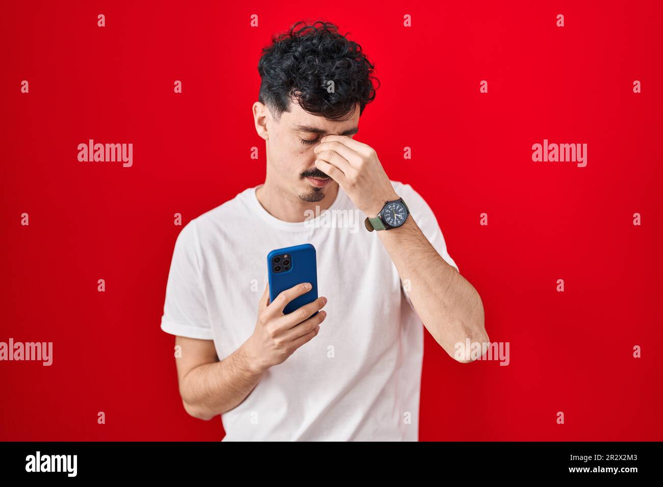 Hispanic man using smartphone over red background tired rubbing nose ...