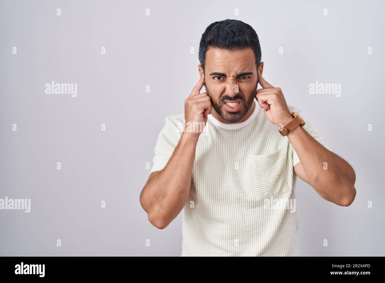 Hispanic man with beard standing over isolated background covering ears ...