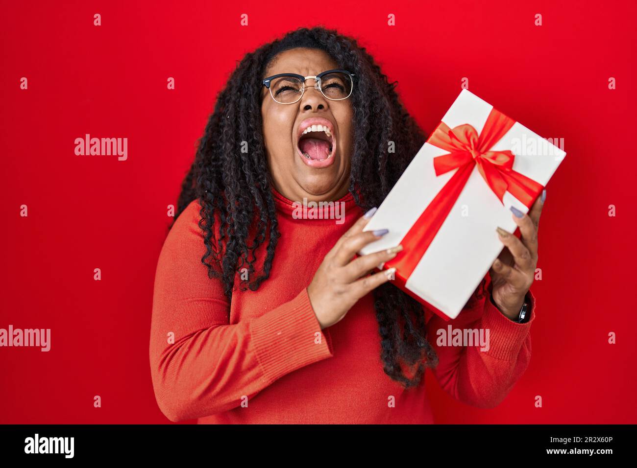 Plus size hispanic woman holding gift angry and mad screaming ...