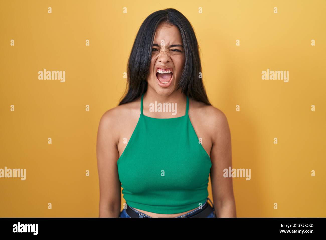 Brunette woman standing over yellow background angry and mad screaming ...