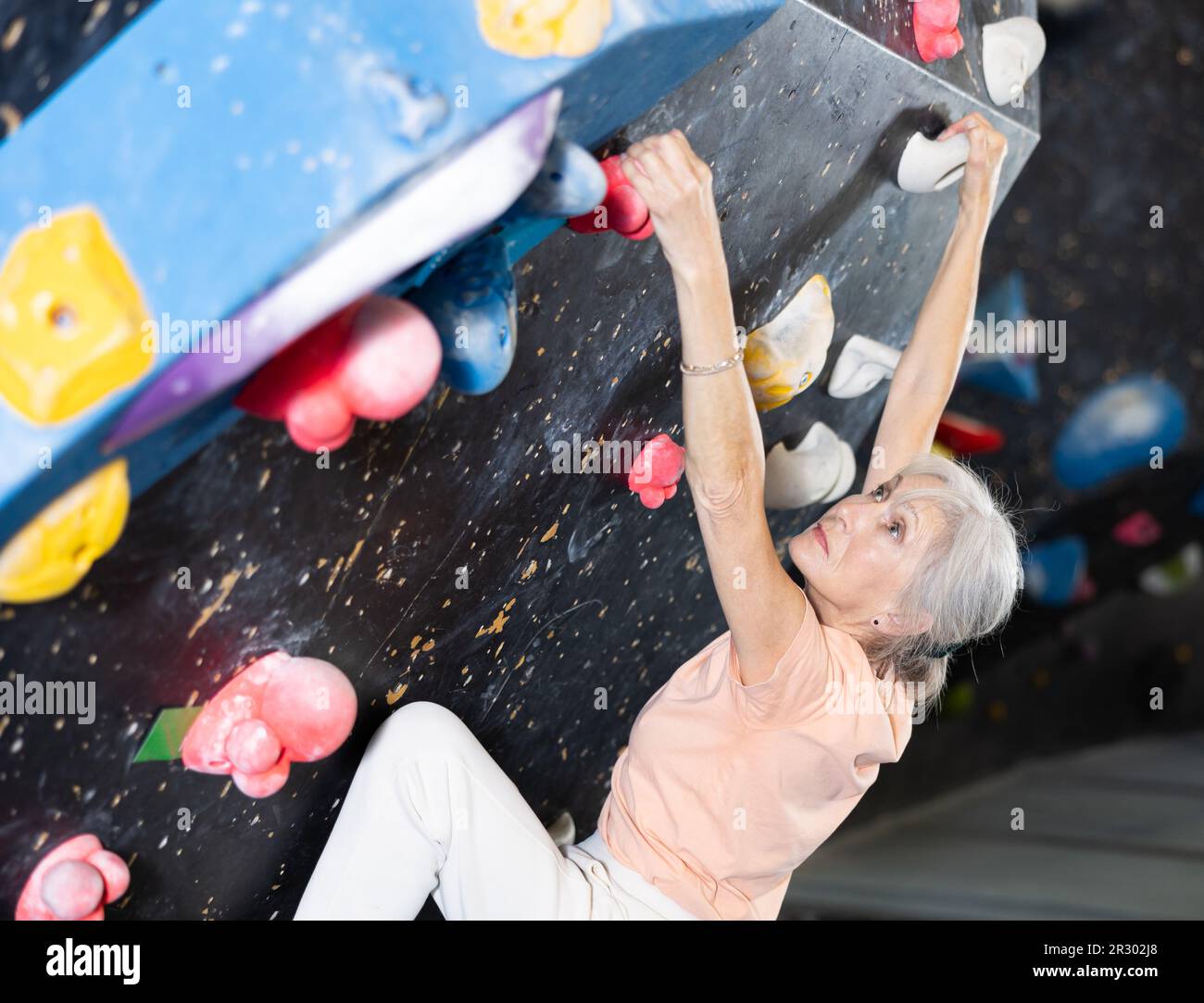 Confident mature woman scaling artificial climbing wall in climbing gym ...