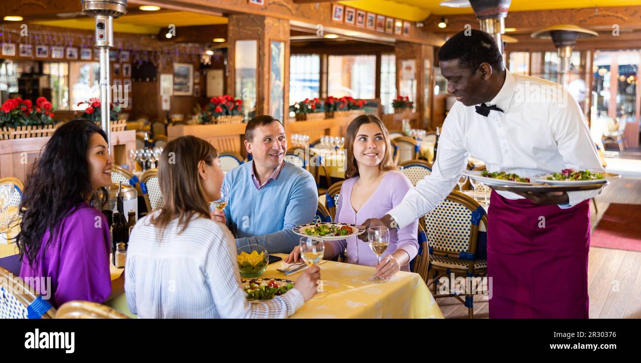 African american waiter serving dishes to cheerful group of adults in ...