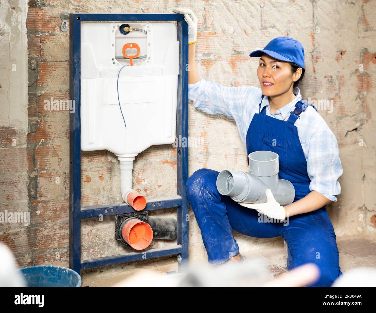 Portrait of female plumber in blue overall holding a plastic sewer pipe ...